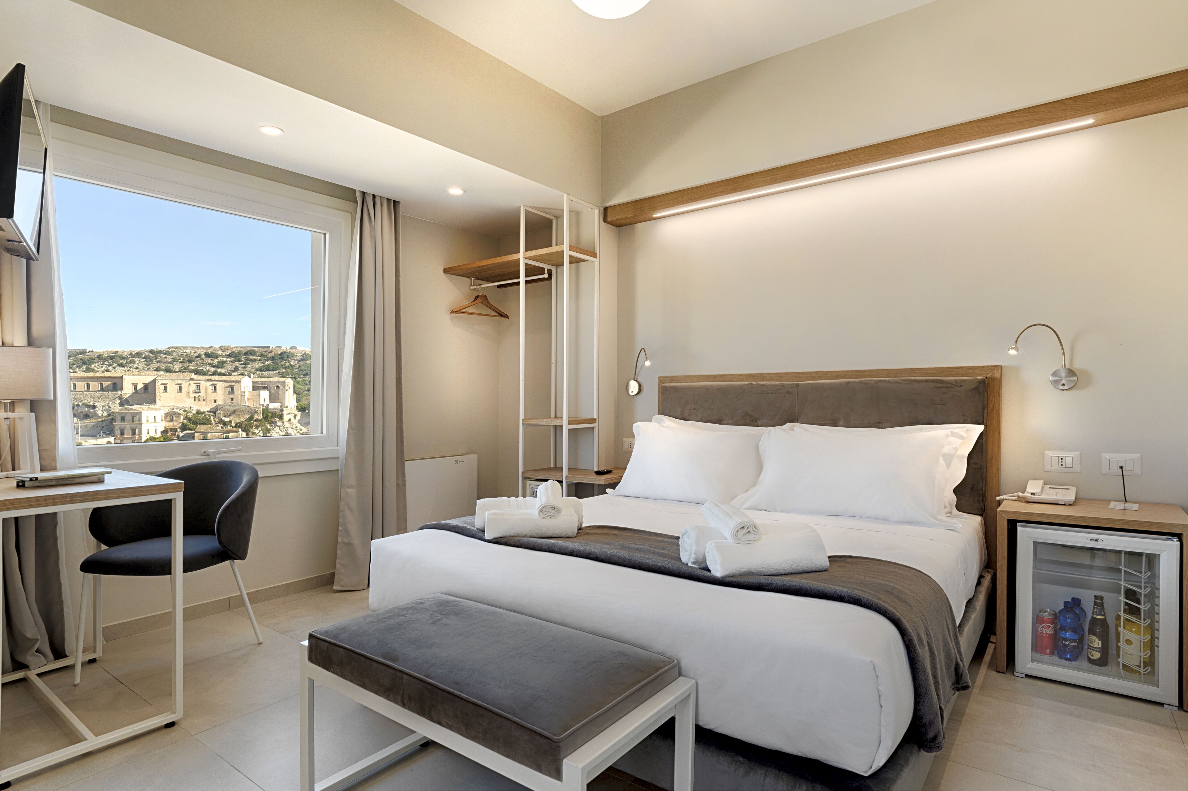 Quarto de hotel moderno com cama, mesa de cabeceira, cadeira e um pequeno frigorífico.