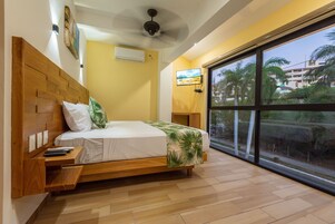 Quarto com cabeceira de madeira, cama com colcha branca e padrão de folhas de palmeira verdes, mesa de cabeceira com controle remoto e uma grande janela com vista para palmeiras e edifícios.