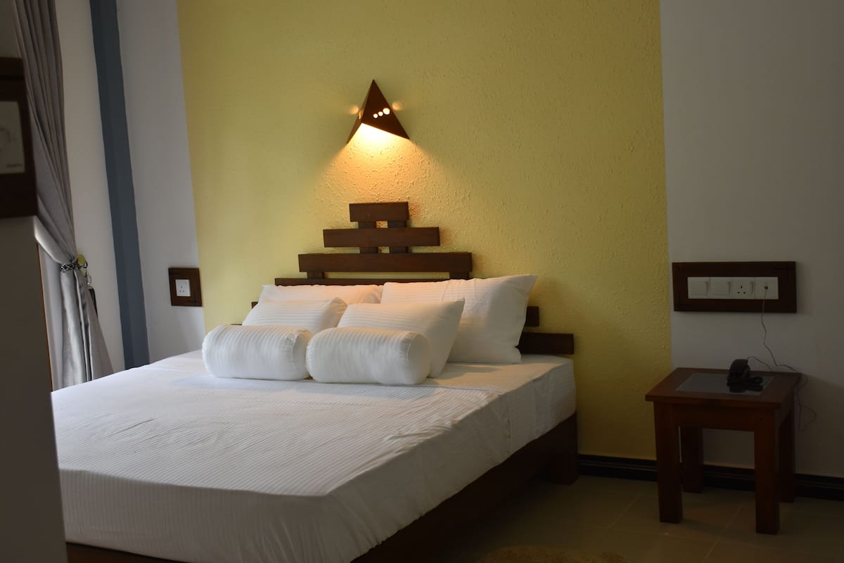 Cama bem arrumada com lençóis e almofadas brancas, mesa de cabeceira de madeira e luminária na parede.