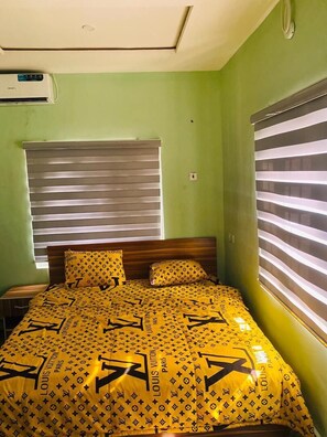 Quarto com uma cama coberta por roupa de cama amarela estampada com padrão Louis Vuitton, duas almofadas, cabeceira de madeira e janela com persianas.