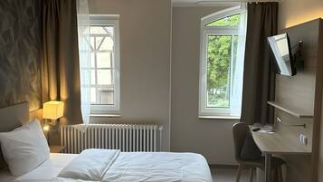 Quarto de hotel com uma cama, uma escrivaninha, uma cadeira, uma televisão e duas janelas com cortinas.