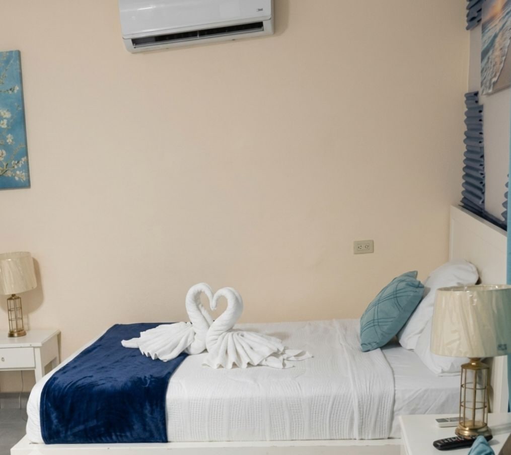 Quarto de hotel com cama, ar-condicionado fixado na parede, luminária de cabeceira e arranjo decorativo de toalhas.