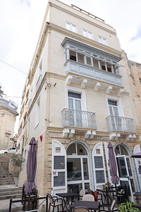 Um edifício de vários andares com uma varanda no segundo andar, área de estar externa com guarda-sóis e uma escadaria para uma entrada superior.