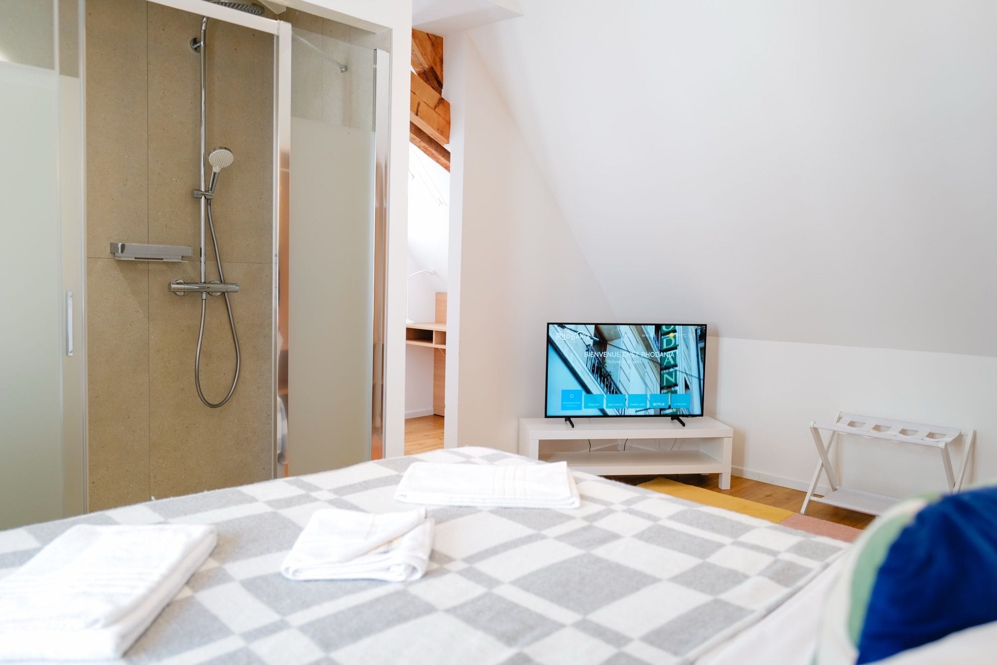 Quarto de hotel com chuveiro, uma cama com colcha xadrez, uma televisão em um suporte e uma parede branca.