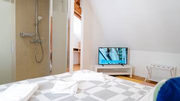 Quarto de hotel com chuveiro, uma cama com colcha xadrez, uma televisão em um suporte e uma parede branca.