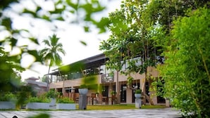 Um edifício moderno com área de estar coberta ao ar livre, rodeado por vegetação e palmeiras.