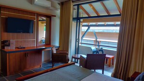 Quarto de hotel com cama, escrivaninha com cadeira, televisão e uma janela grande com cortinas.