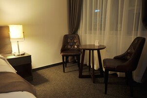Quarto de hotel com cama, mesa de cabeceira com abajur, duas poltronas de couro, uma pequena mesa e uma janela com cortinas.