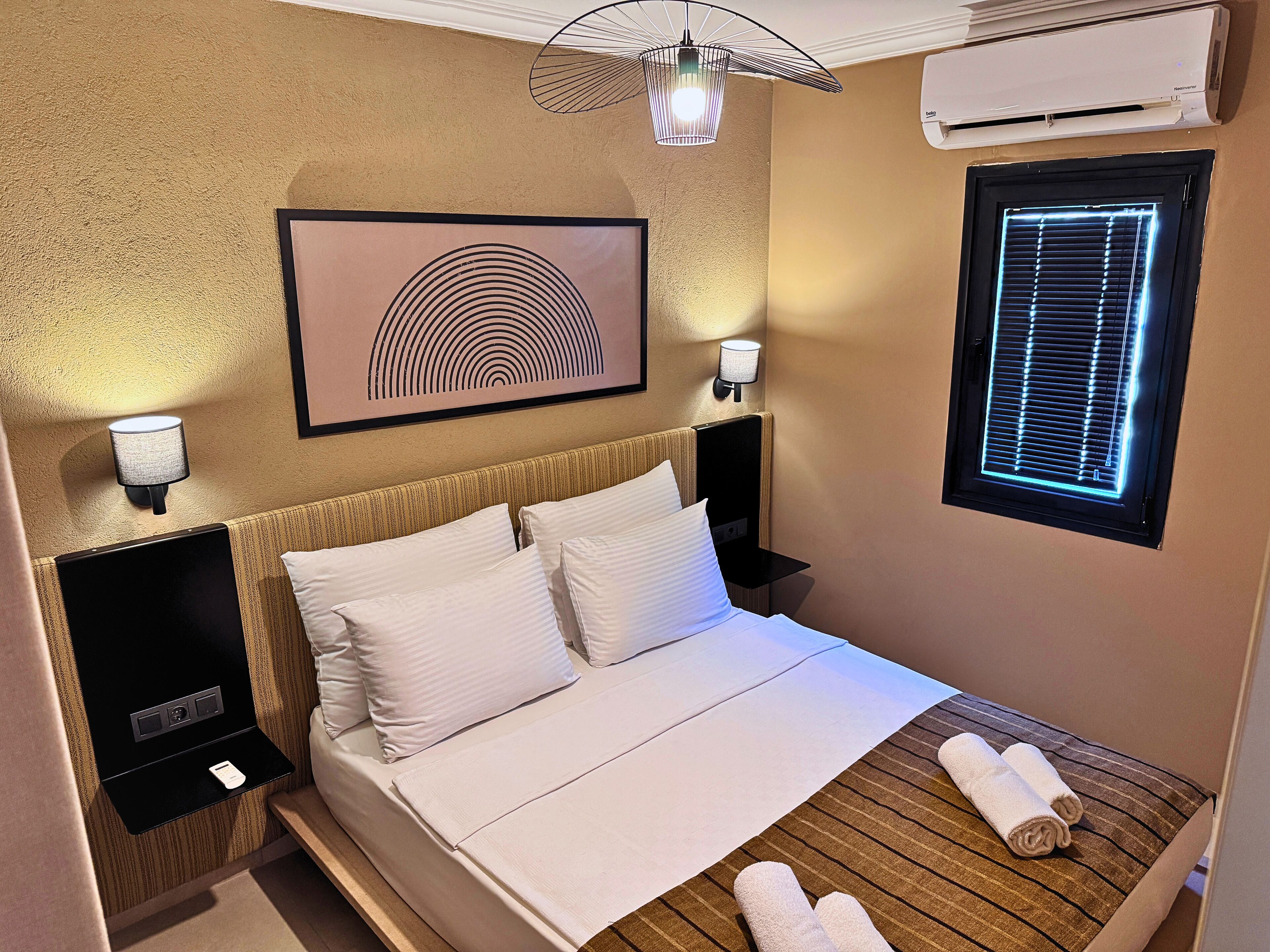 Quarto de hotel com cama, mesas de cabeceira, luminária na parede, ar condicionado e janela com persianas.