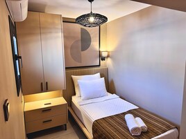 Quarto de hotel compacto com cama, mesa de cabeceira, guarda-roupa e luminária fixada na parede.