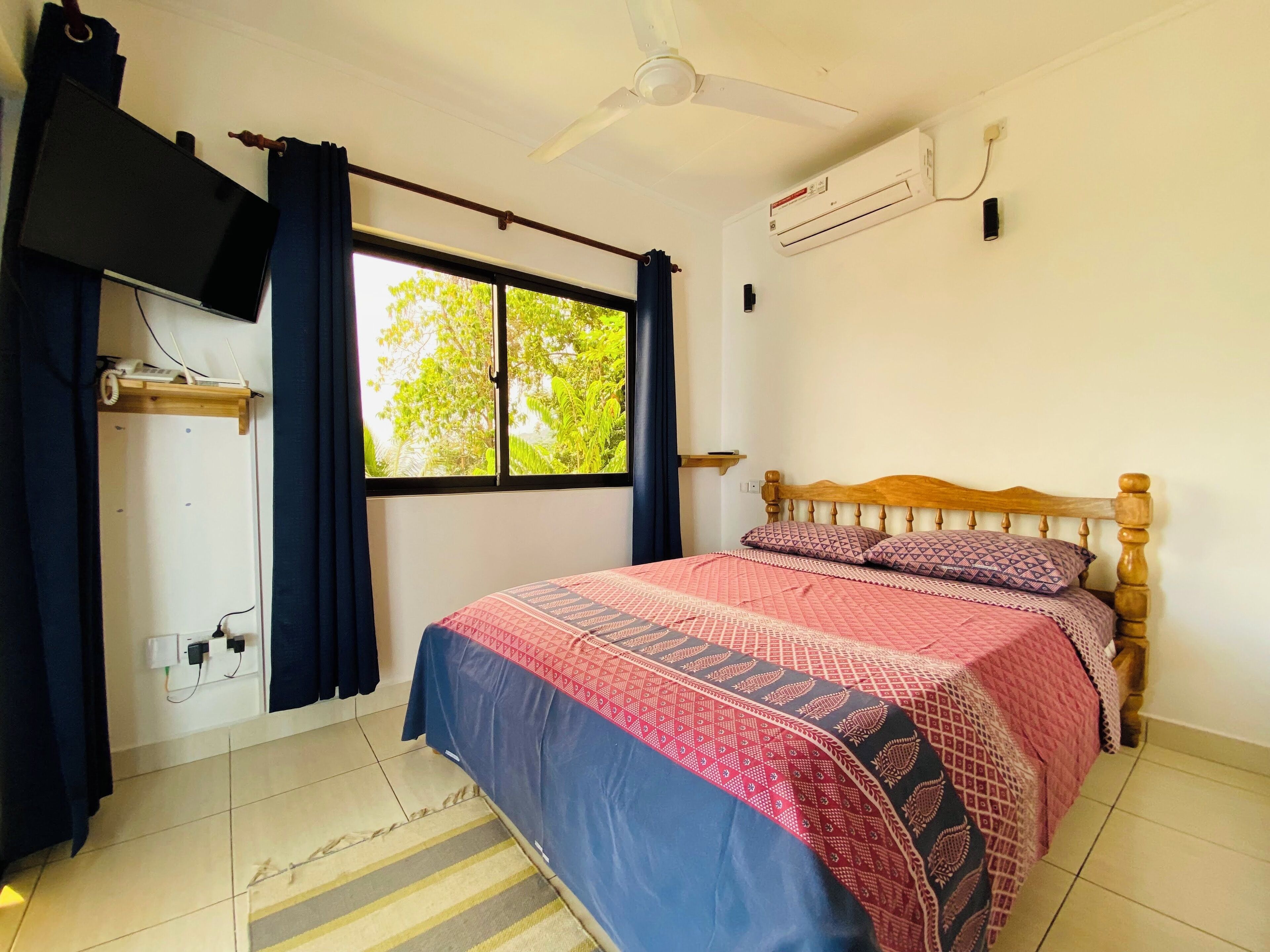 Quarto com cama, ventilador de teto, ar condicionado, janela com cortinas e TV fixada na parede.