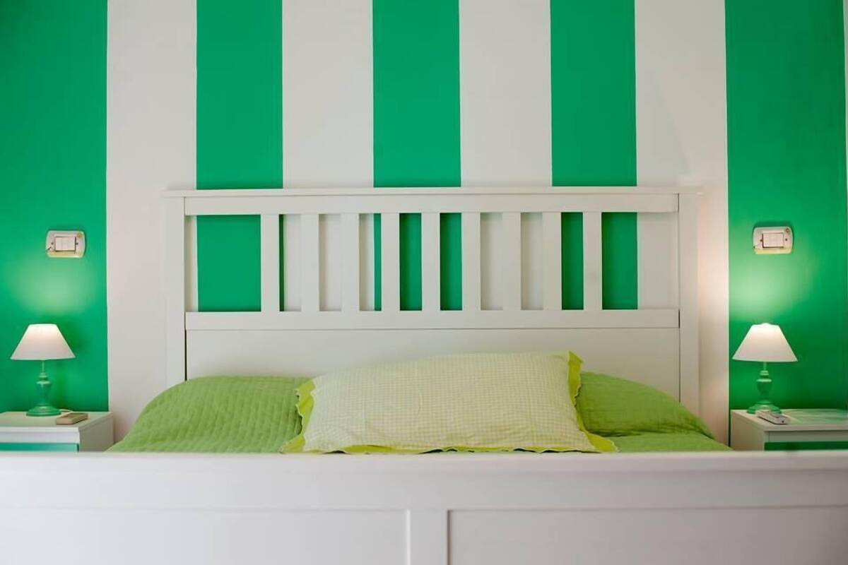 Uma cama com cabeceira branca, paredes verdes e duas luminárias de cabeceira.