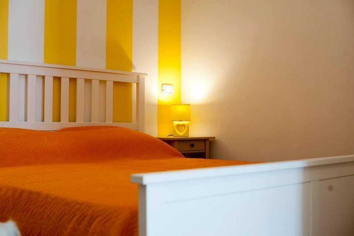 Uma cama com colcha laranja, cabeceira branca e mesa de cabeceira com abajur amarelo.