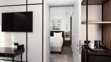Quarto de hotel moderno com televisão emoldurada a preto, cama com roupa de cama branca, mesa de cabeceira com chaleira e máquina de café.