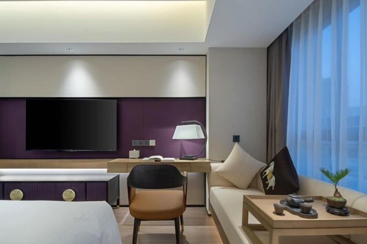 Quarto de hotel moderno com uma televisão de tela plana grande, um sofá, uma cadeira, uma escrivaninha e uma mesinha de centro com objetos decorativos.