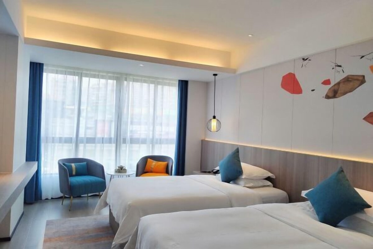 Quarto de hotel com duas camas, uma escrivaninha, uma cadeira e uma janela grande com cortinas.