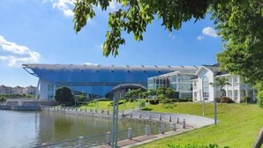 Um edifício moderno com um telhado curvo distinto, um lago e uma área gramada bem cuidada com um caminho.