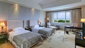 Quarto de hotel com duas camas, uma mesa, uma cadeira, uma luminária e vista para o mar.
