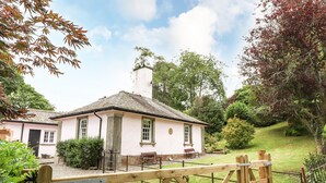 Uma casa de campo branca com telhado de palha, um portão de madeira e um jardim com um banco e uma mesa pequena.