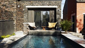 Piscina com duas espreguiçadeiras e uma pequena mesa, cercada por muros de pedra e um deck de madeira.