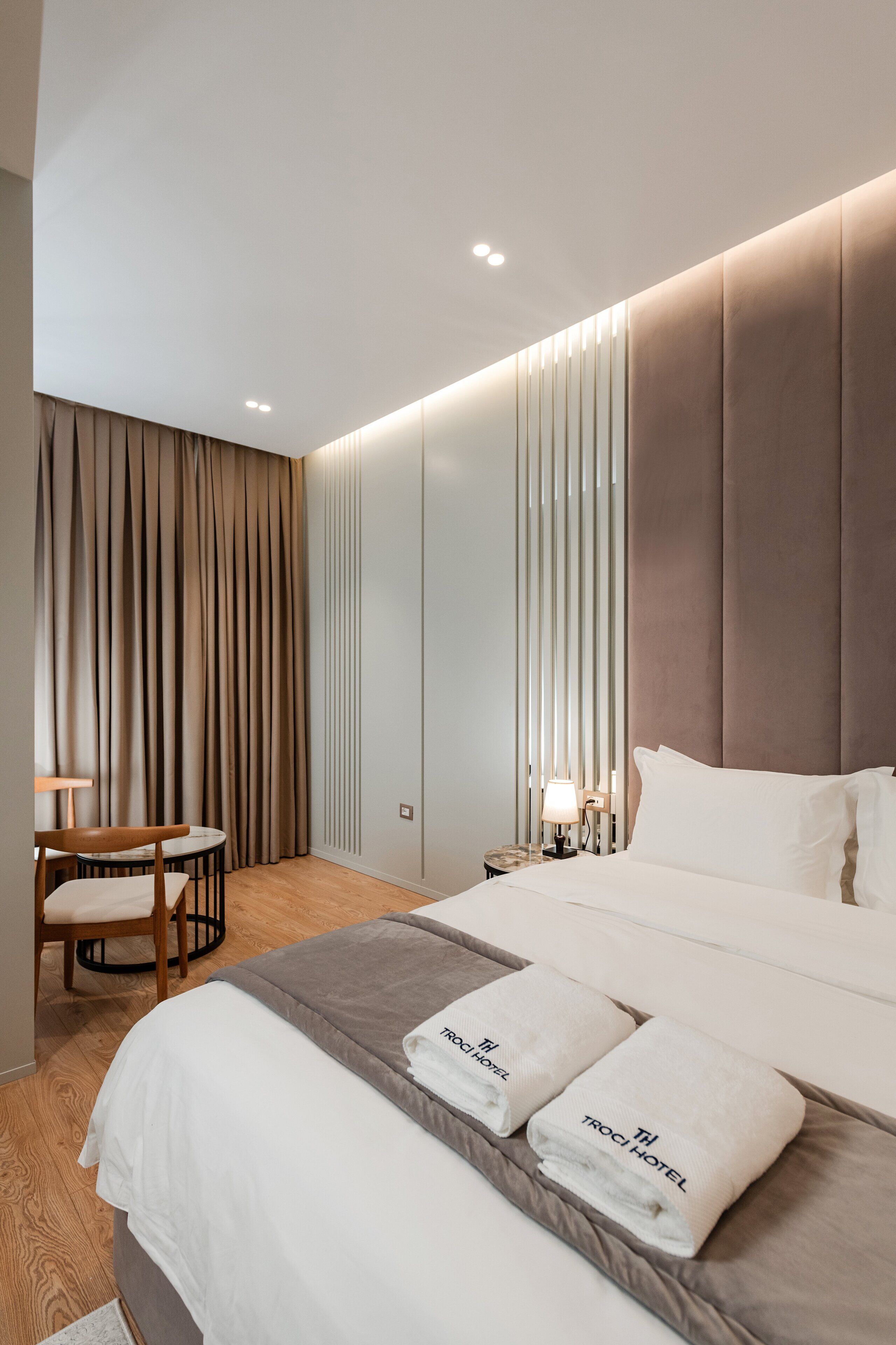 Quarto de hotel moderno com uma cama grande, mesa de cabeceira com luminárias, uma cadeira e vista para as cortinas.