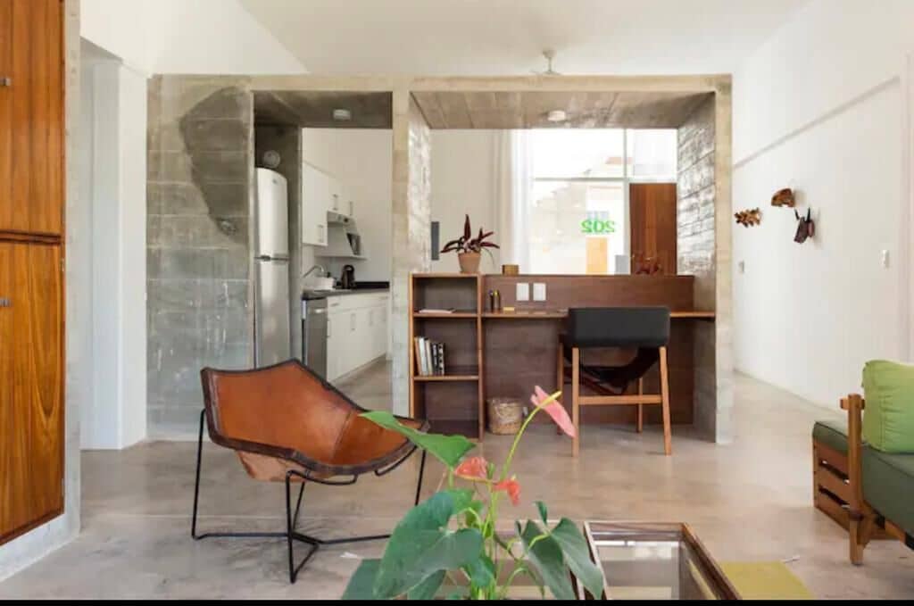 Uma sala de estar moderna com uma poltrona de couro marrom, um sofá verde e uma mesa de centro. Há uma área de cozinha com uma geladeira e uma pia ao fundo.