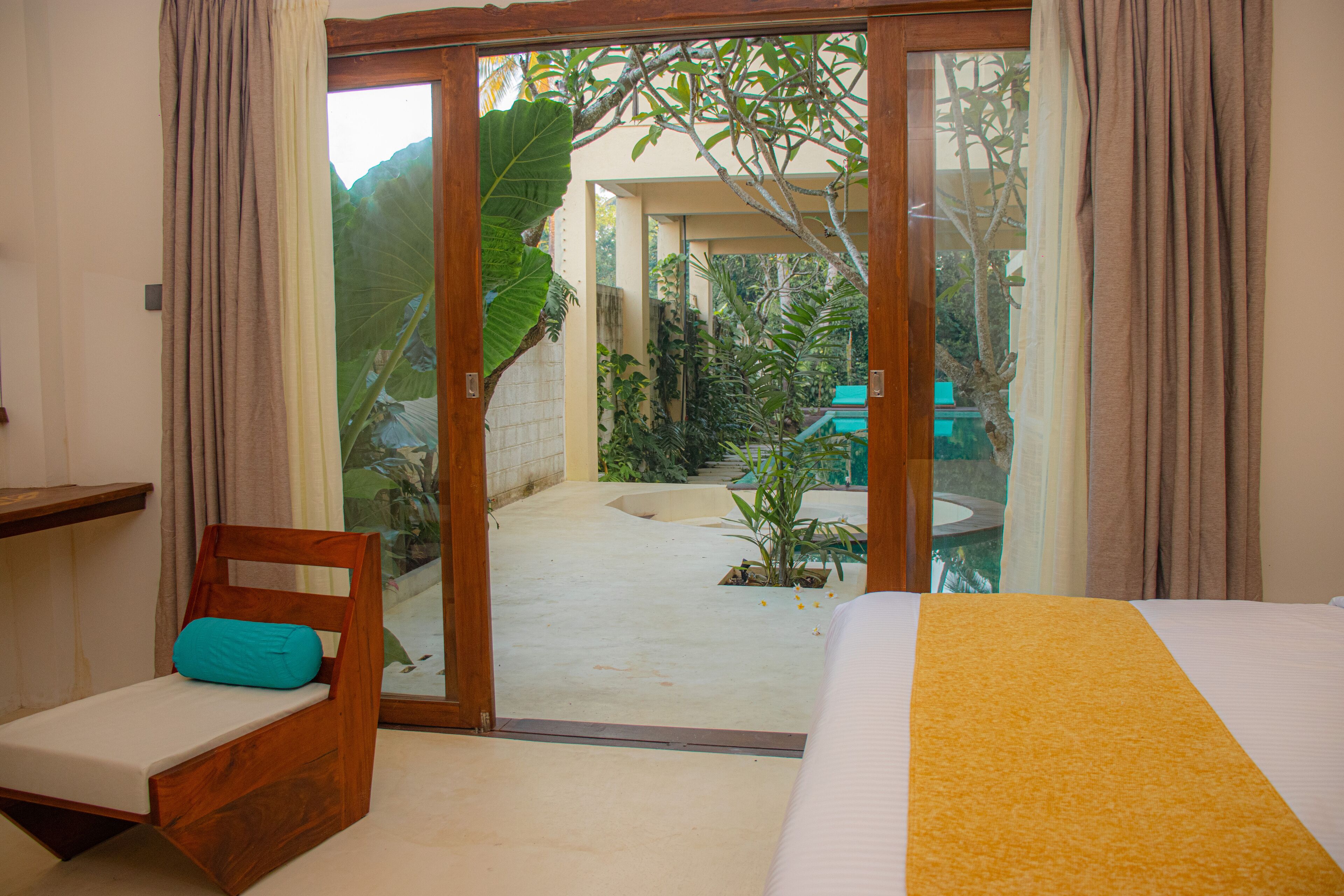 Um quarto com uma cama, uma cadeira de madeira com almofada e uma porta de vidro deslizante para um alpendre com plantas e uma mesa pequena.