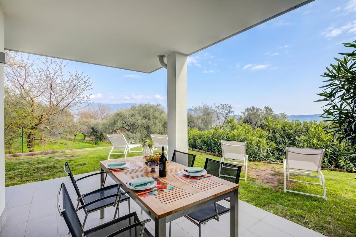 Uma área de refeições ao ar livre com uma mesa posta para quatro pessoas, rodeada por cadeiras, e vista para uma paisagem gramada com árvores e colinas.