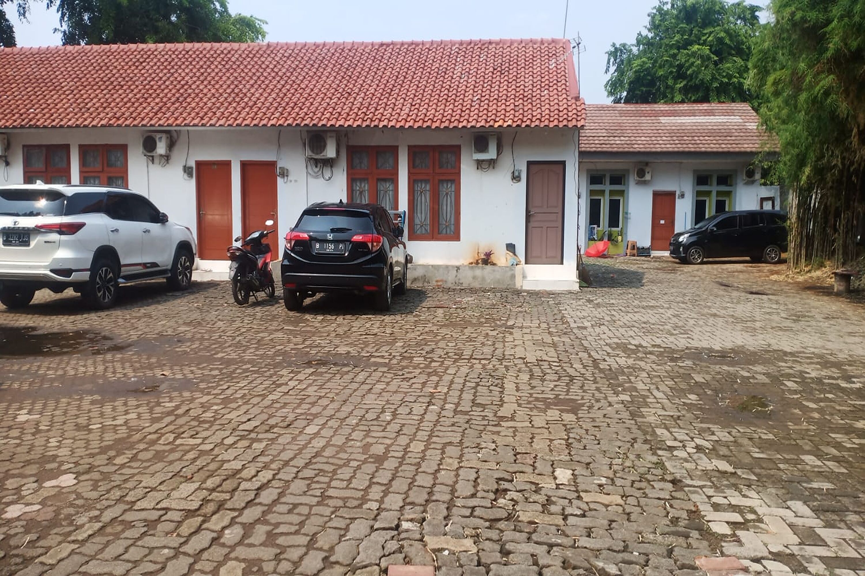 Um edifício com telhado de telhas vermelhas e paredes brancas, com entrada por um caminho pavimentado e veículos estacionados.