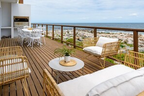 Um deck de madeira com móveis de exterior, incluindo cadeiras de vime e uma mesa, com vista para um litoral rochoso.