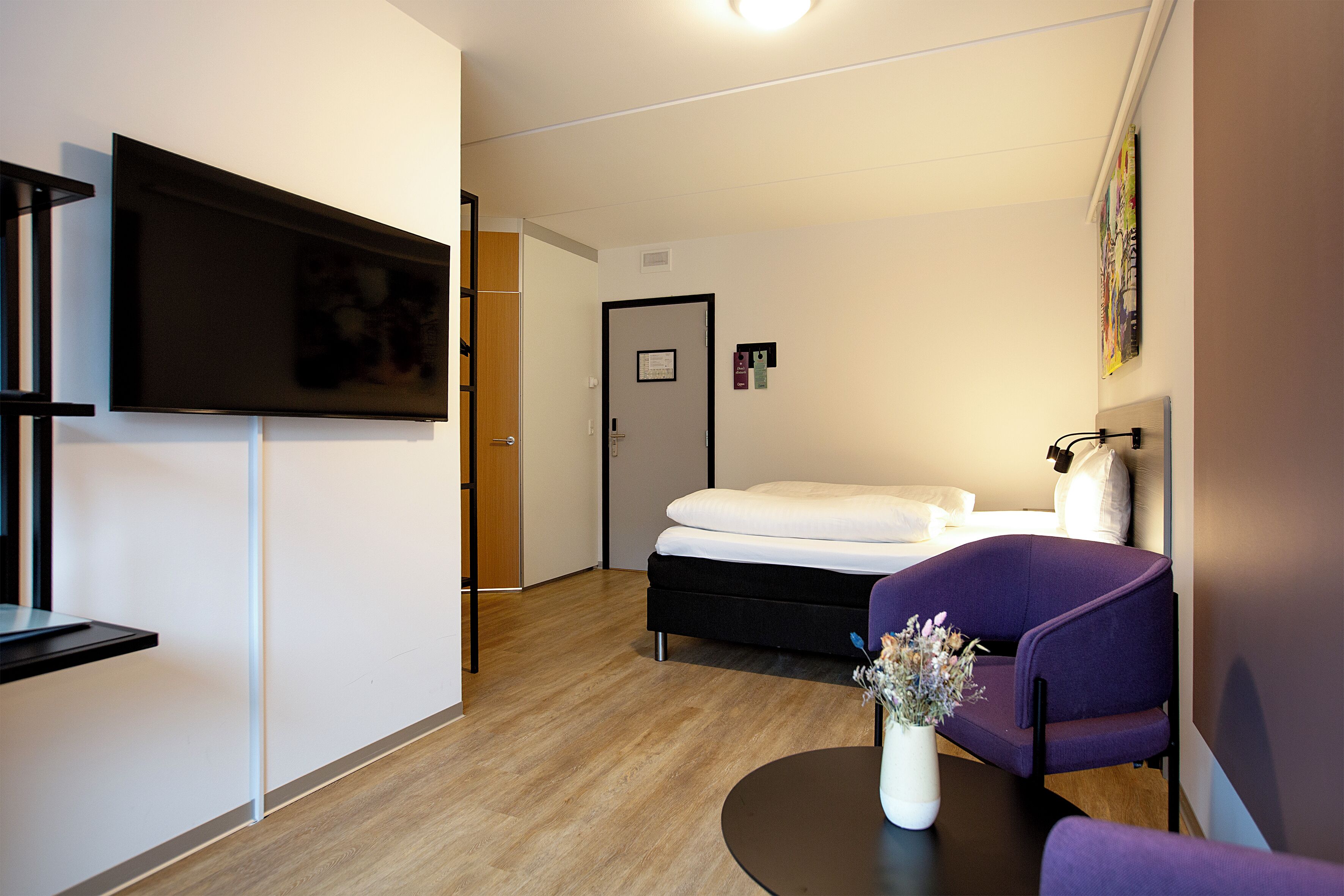 Quarto de hotel com cama, televisão de tela plana na parede, poltrona roxa, mesa redonda pequena com um vaso de flores e piso de madeira.