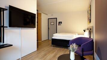 Quarto de hotel com cama, televisão de tela plana na parede, poltrona roxa, mesa redonda pequena com um vaso de flores e piso de madeira.