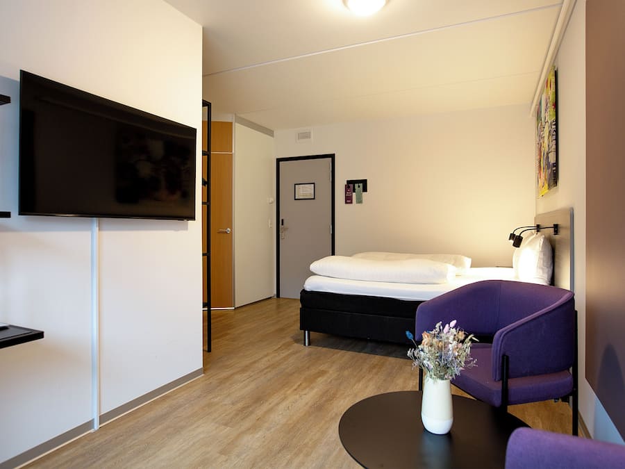 Quarto de hotel com cama, televisão de tela plana na parede, poltrona roxa, mesa redonda pequena com um vaso de flores e piso de madeira.