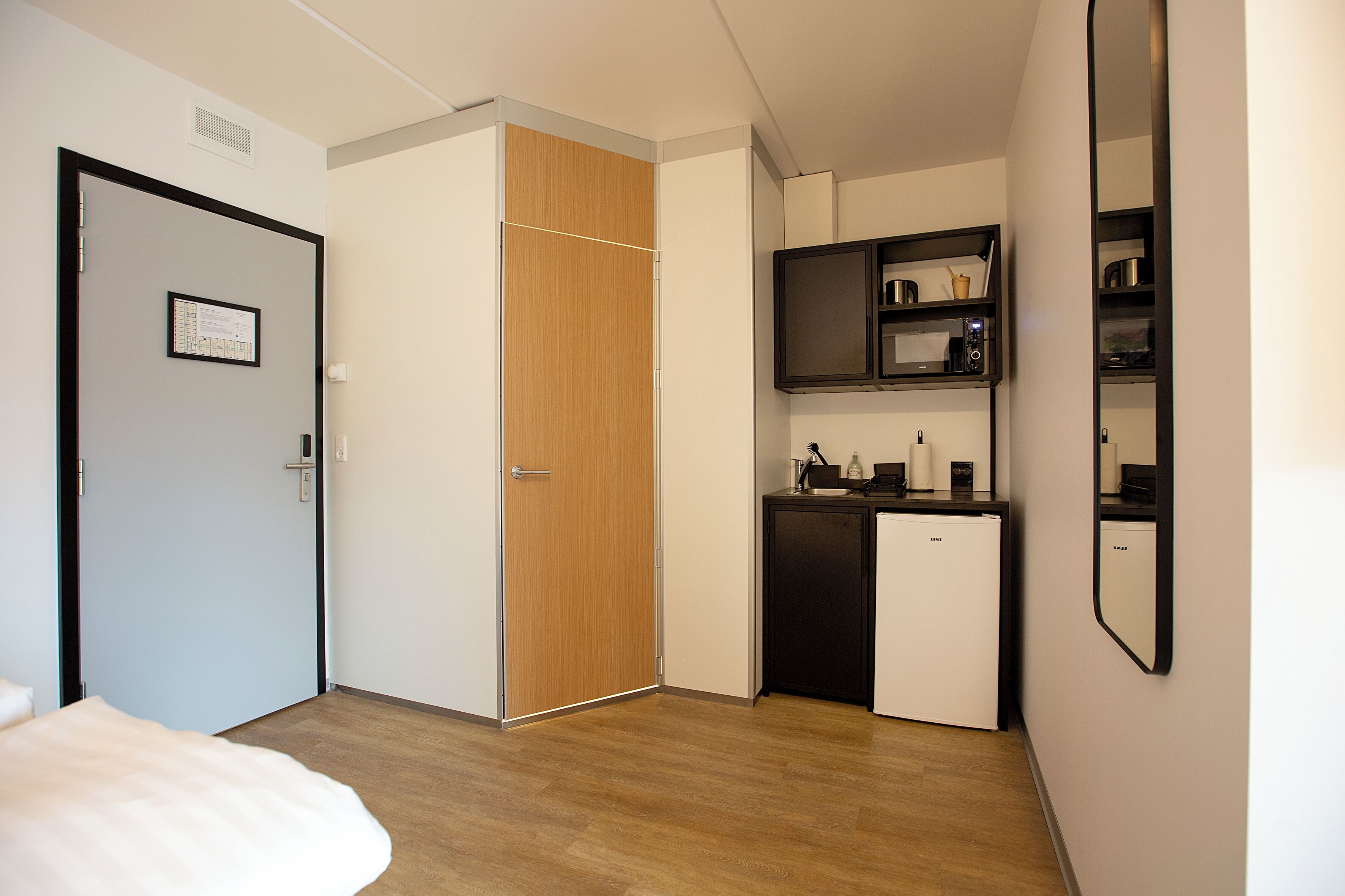Quarto de hotel pequeno e funcional, com uma cama, guarda-roupa de madeira, kitchenette e espelho embutido.