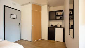 Quarto de hotel pequeno e funcional, com uma cama, guarda-roupa de madeira, kitchenette e espelho embutido.