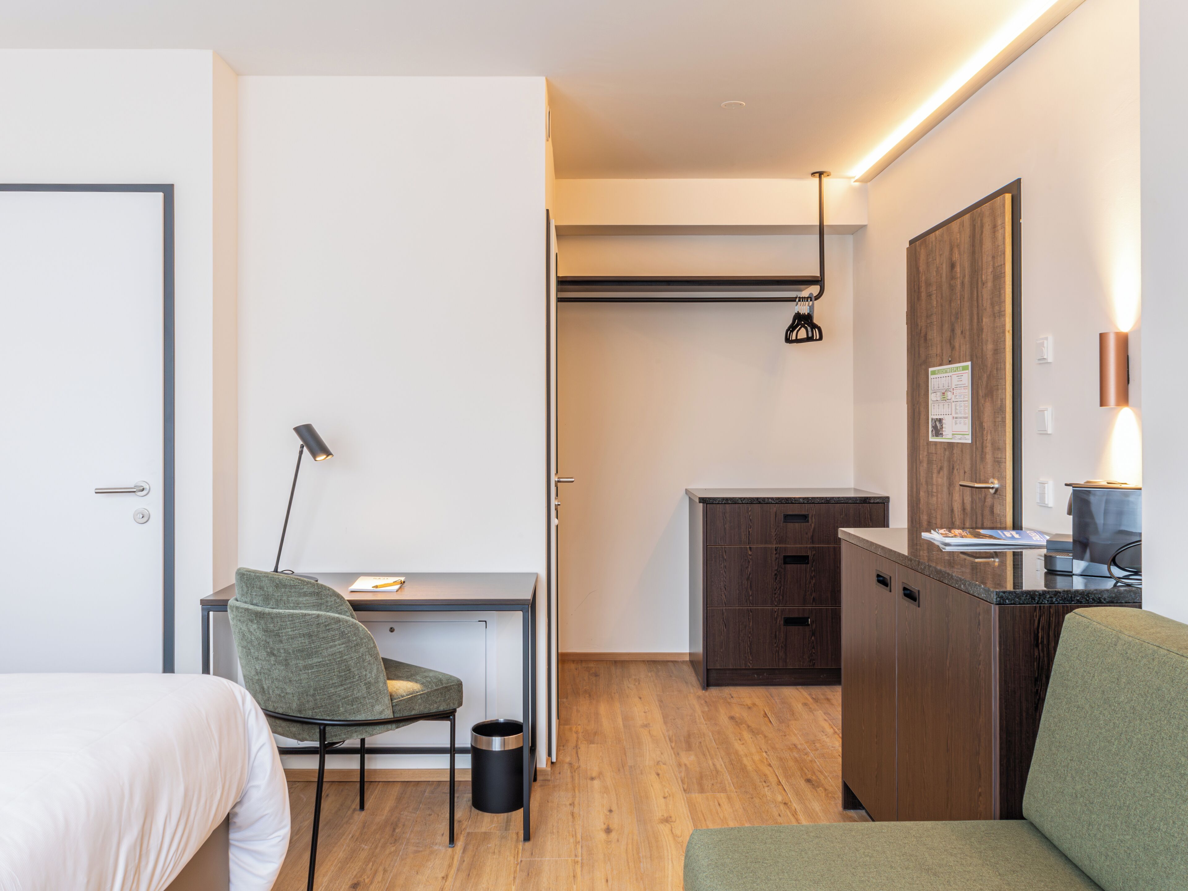 Quarto de hotel compacto com cama, escrivaninha, cadeira, pequena cozinha e armário.