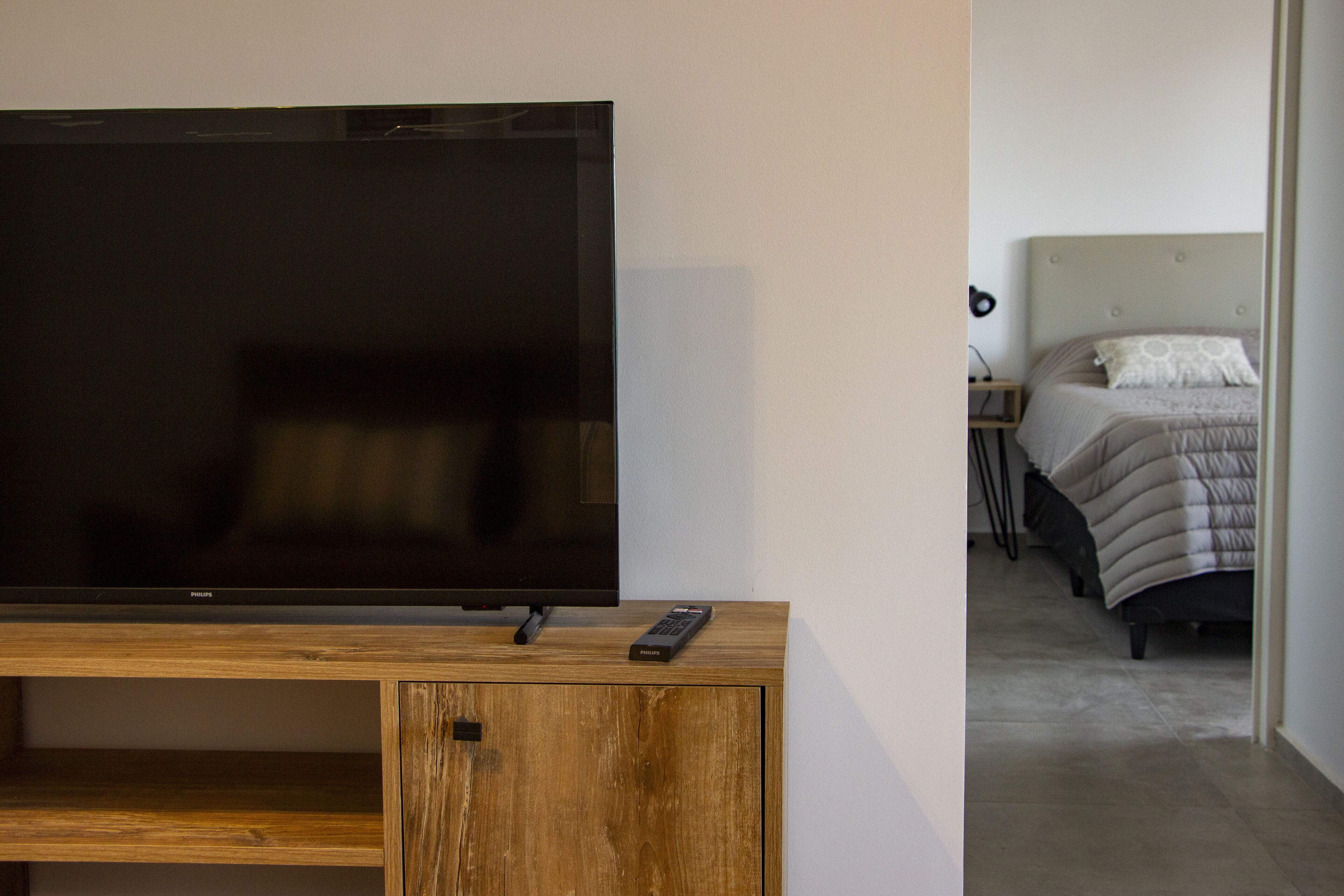 Televisão sobre suporte de madeira com um controle remoto ao lado, uma cama com cabeceira e travesseiros, e uma mesa de cabeceira com uma luminária.