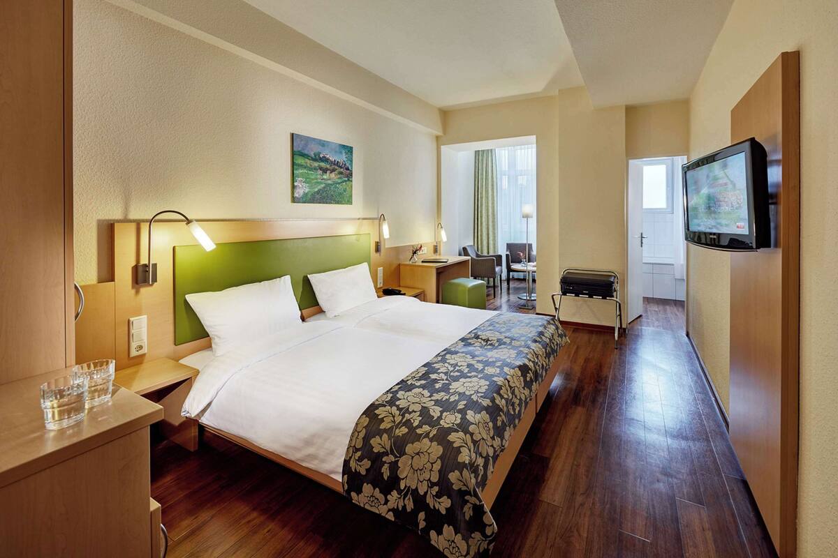 Quarto de hotel com cama, mesas de cabeceira, escrivaninha, cadeira, televisão e um quadro na parede.