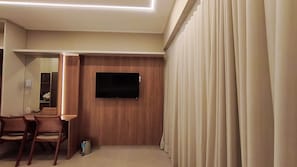 Quarto de hotel com parede de madeira, TV de tela plana fixada na parede, mesa de jantar com cadeiras e cortina na janela.