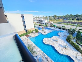 Piscina com design ondulado, cercada por espreguiçadeiras e guarda-sóis, com vista para um edifício e áreas verdes ao fundo.