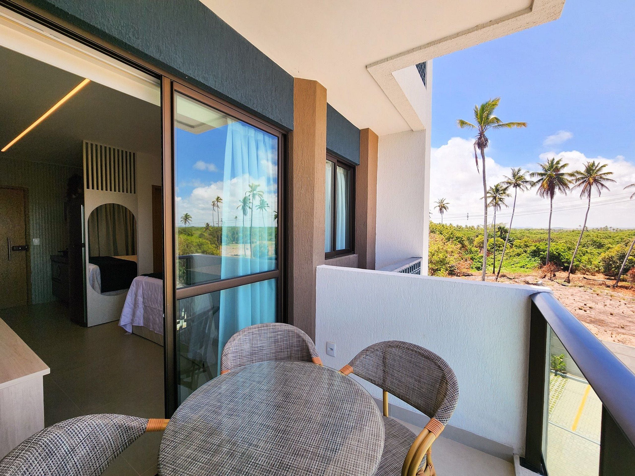 Uma varanda com vista para palmeiras e uma praia, uma mesa redonda com cadeiras, e uma porta de vidro que dá para um quarto com uma cama e uma mesinha.