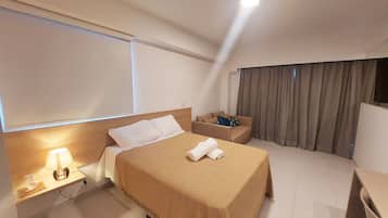 Quarto de hotel com cama, mesa de cabeceira com luminária, sofá e uma grande janela com cortinas.