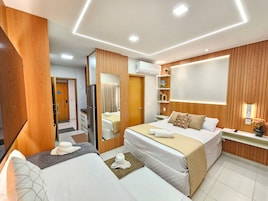 Quarto de hotel com duas camas, cozinha compacta, banheiro e parede de destaque em madeira.