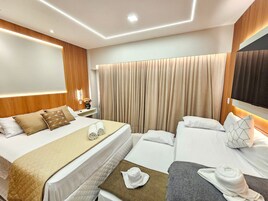 Quarto de hotel com uma cama grande, uma cama menor, uma escrivaninha e uma janela ampla com cortinas.