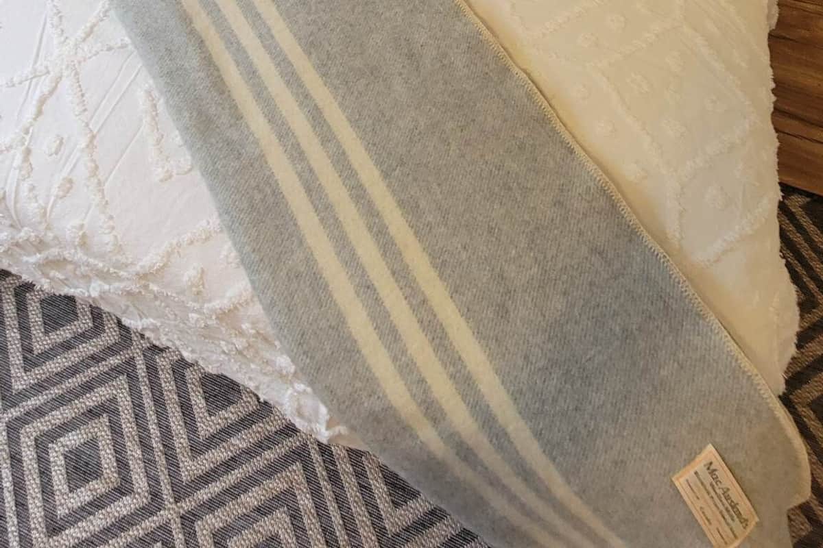 Cama bem arrumada com edredom branco, manta listrada cinza e branca, e travesseiro decorativo com desenho de pássaro.