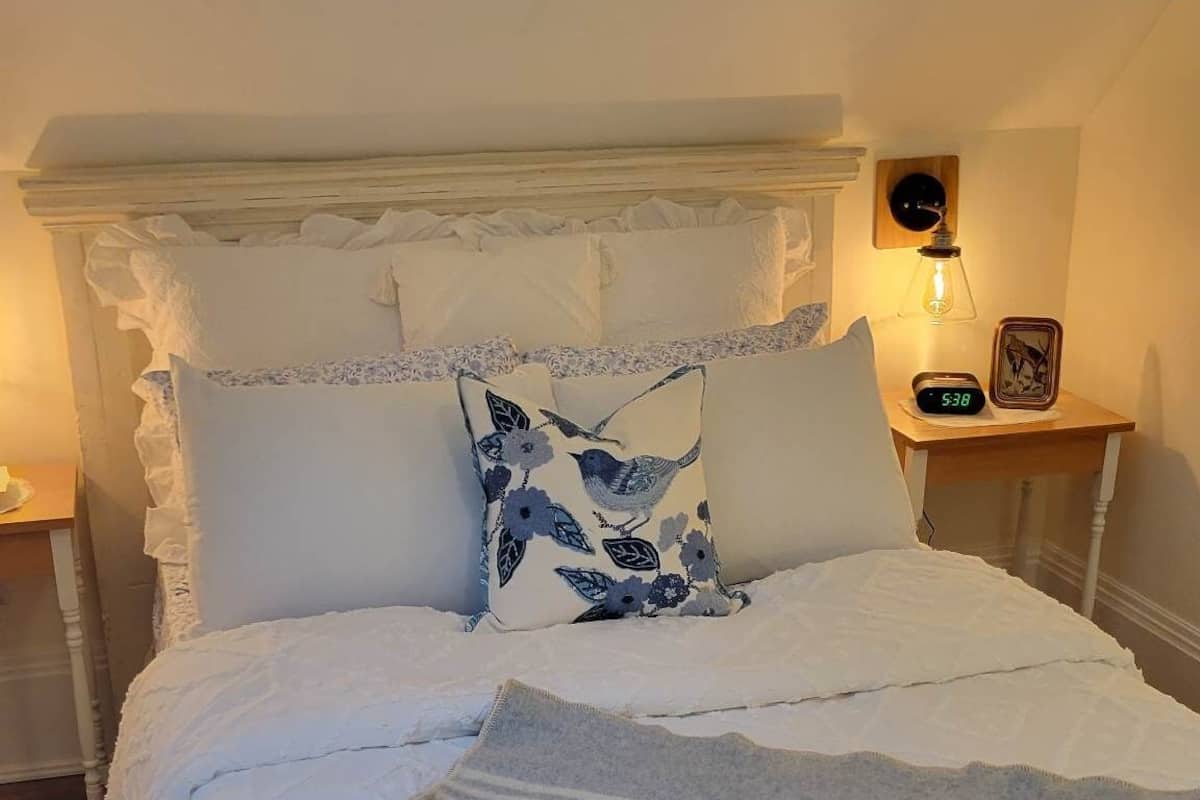 Cama bem arrumada com lençóis brancos e almofada floral azul, mesa de cabeceira com um relógio, e tapete estampado.