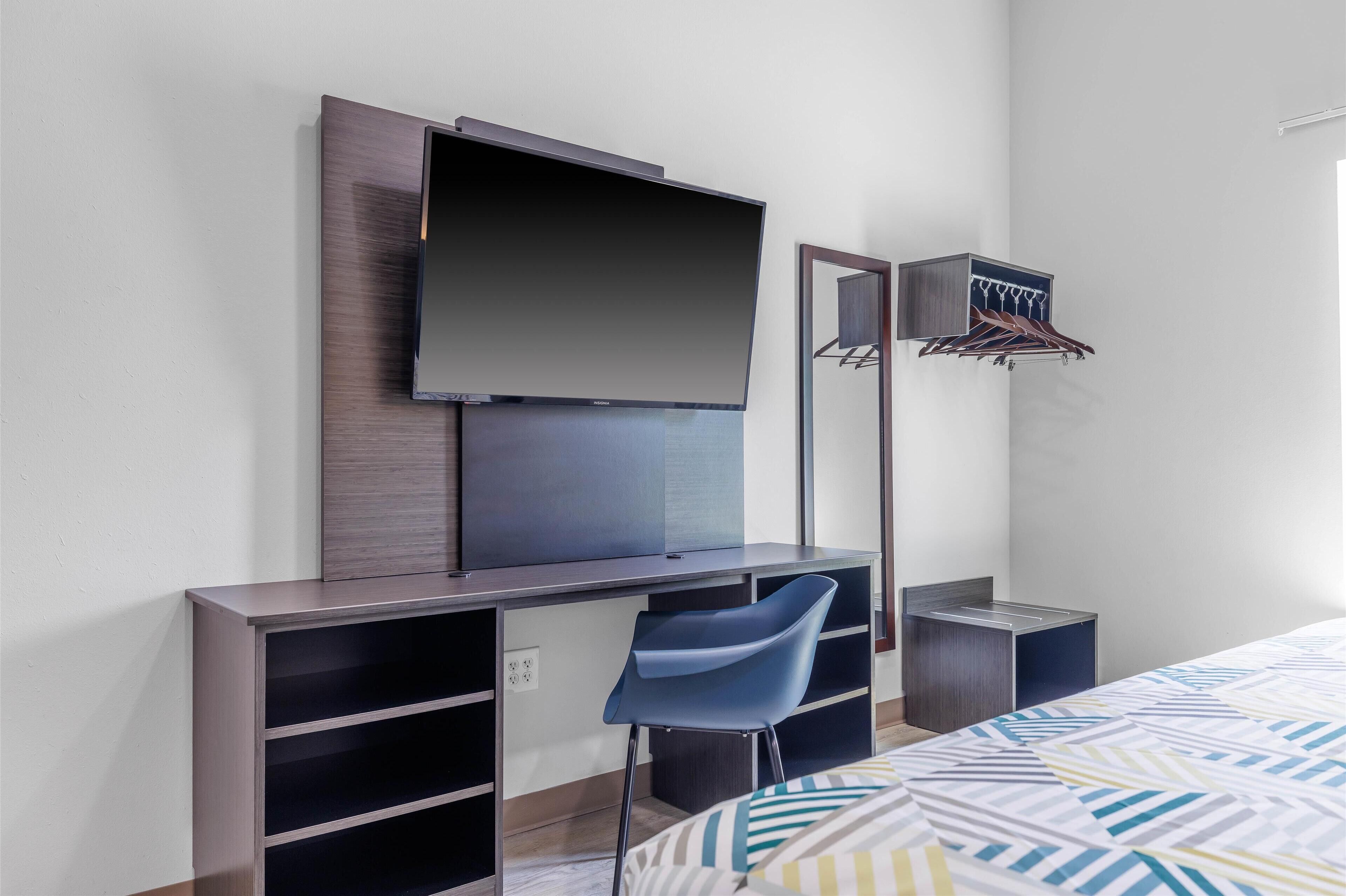 Quarto de hotel moderno com televisão de tela plana na parede, uma escrivaninha com cadeira, uma cama com roupa de cama estampada e um espelho com suporte para pendurar roupas.