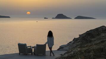 Uma pessoa caminhando por uma costa rochosa, com vista para o mar e ilhas distantes ao pôr do sol.