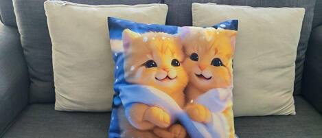 Um sofá cinzento com duas almofadas bege e uma almofada decorativa com gatinhos.