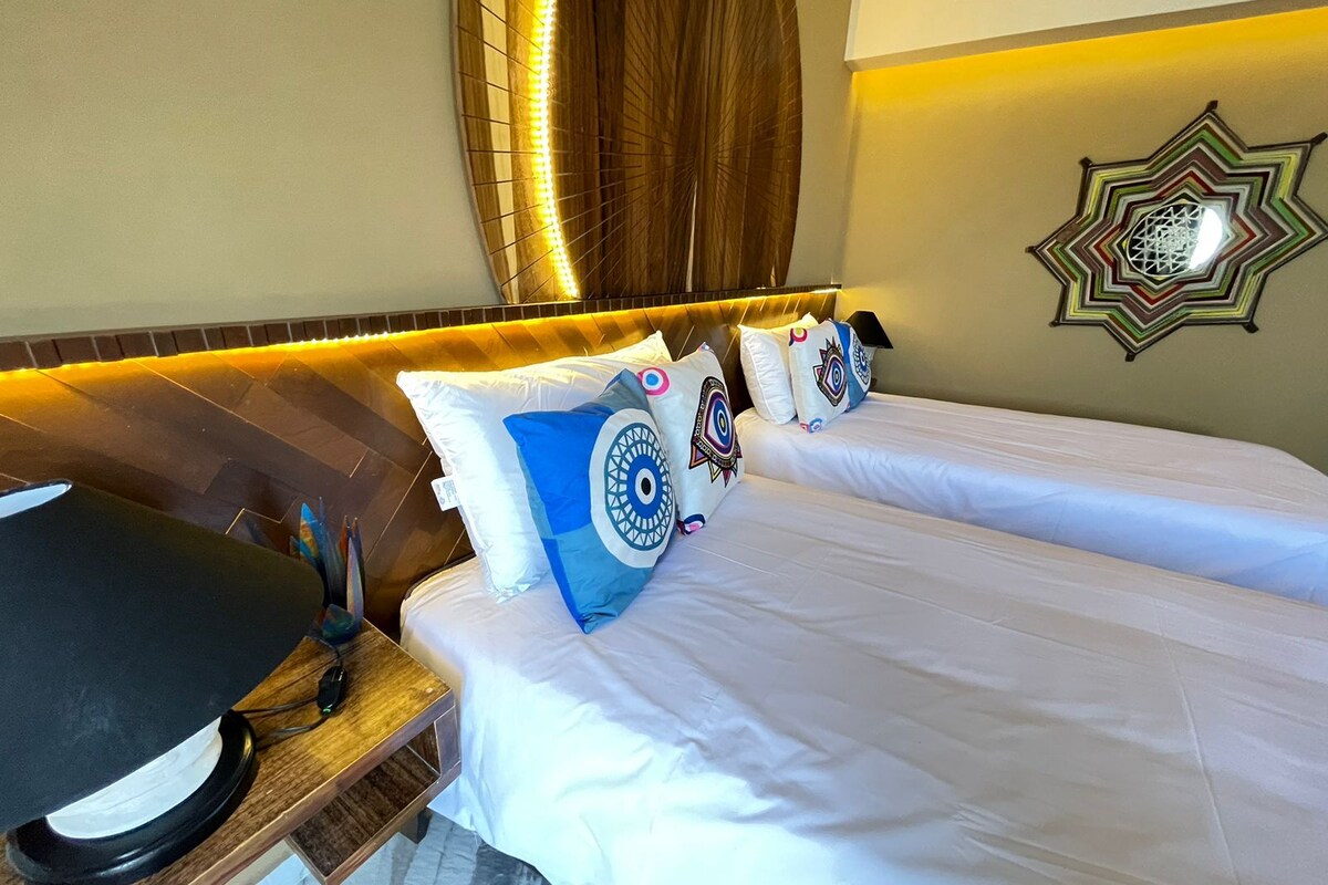 Quarto de hotel com cama, mesa de cabeceira, luminária, almofadas decorativas e uma obra de arte na parede.
